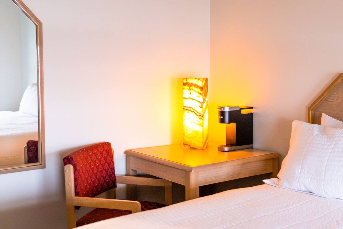Quarto de hotel com cama, mesa de cabeceira com abajur, cadeira e espelho.