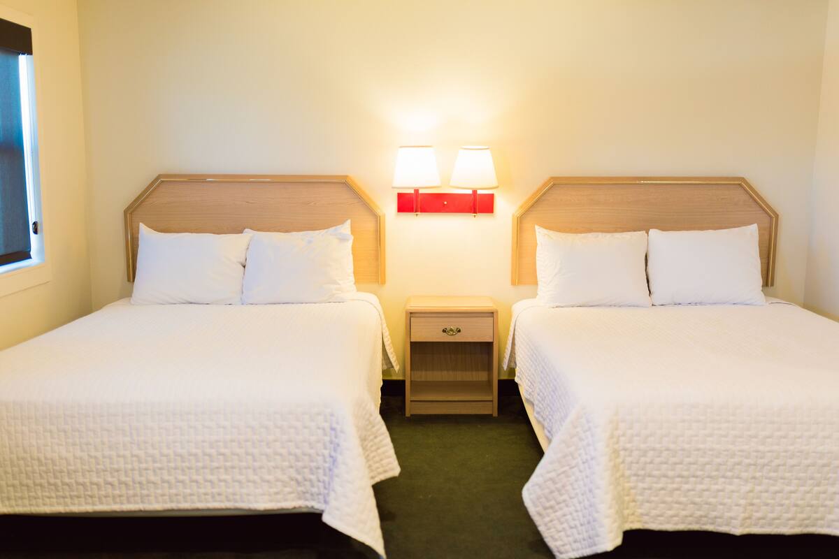 Quarto de hotel com duas camas, uma mesa de cabeceira com abajur e uma janela.