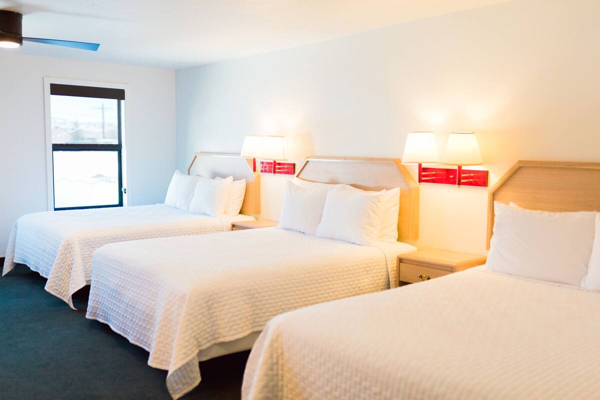 Quarto de hotel com três camas de solteiro, cada uma com colcha e travesseiros brancos, e luminárias de cabeceira com base vermelha.