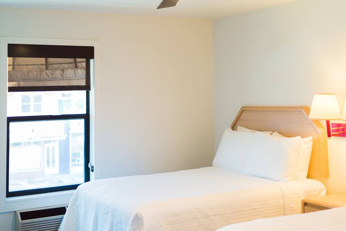 Quarto de hotel com duas camas, um ventilador de teto, janela com vista e uma luminária de cabeceira.