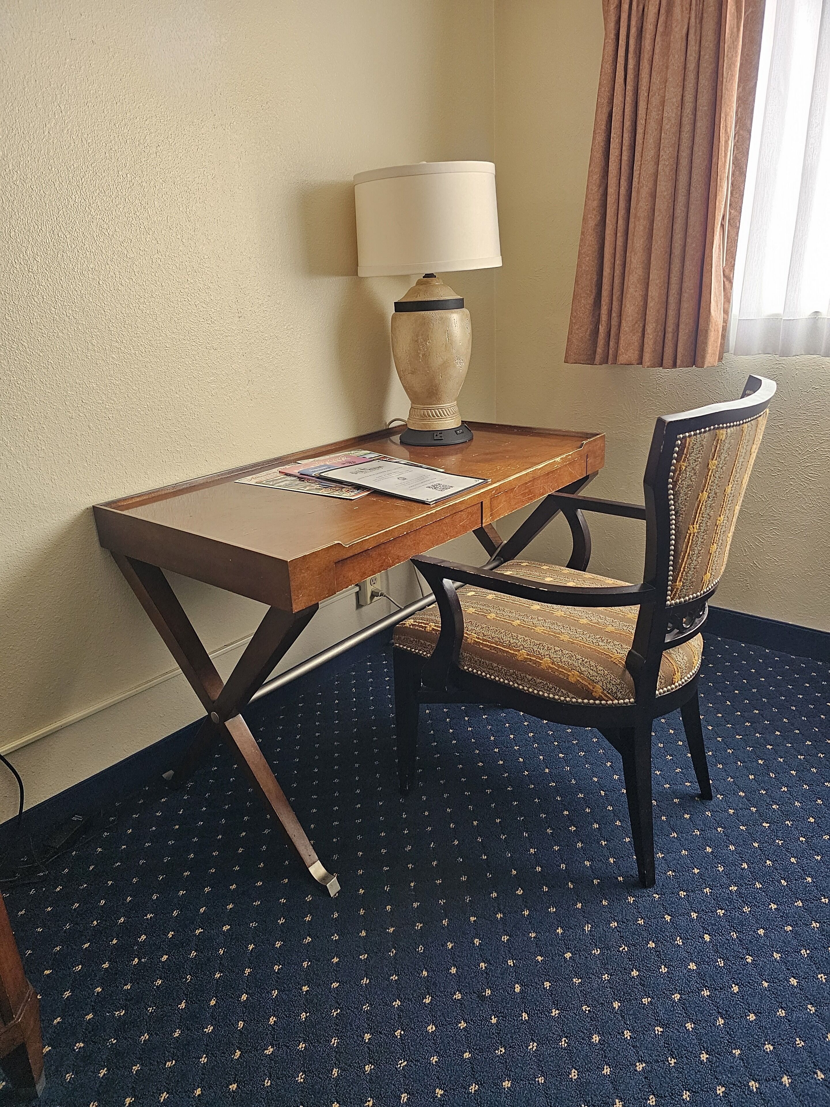 Quarto de hotel com escrivaninha de madeira, uma cadeira, luminária e janela com cortinas.