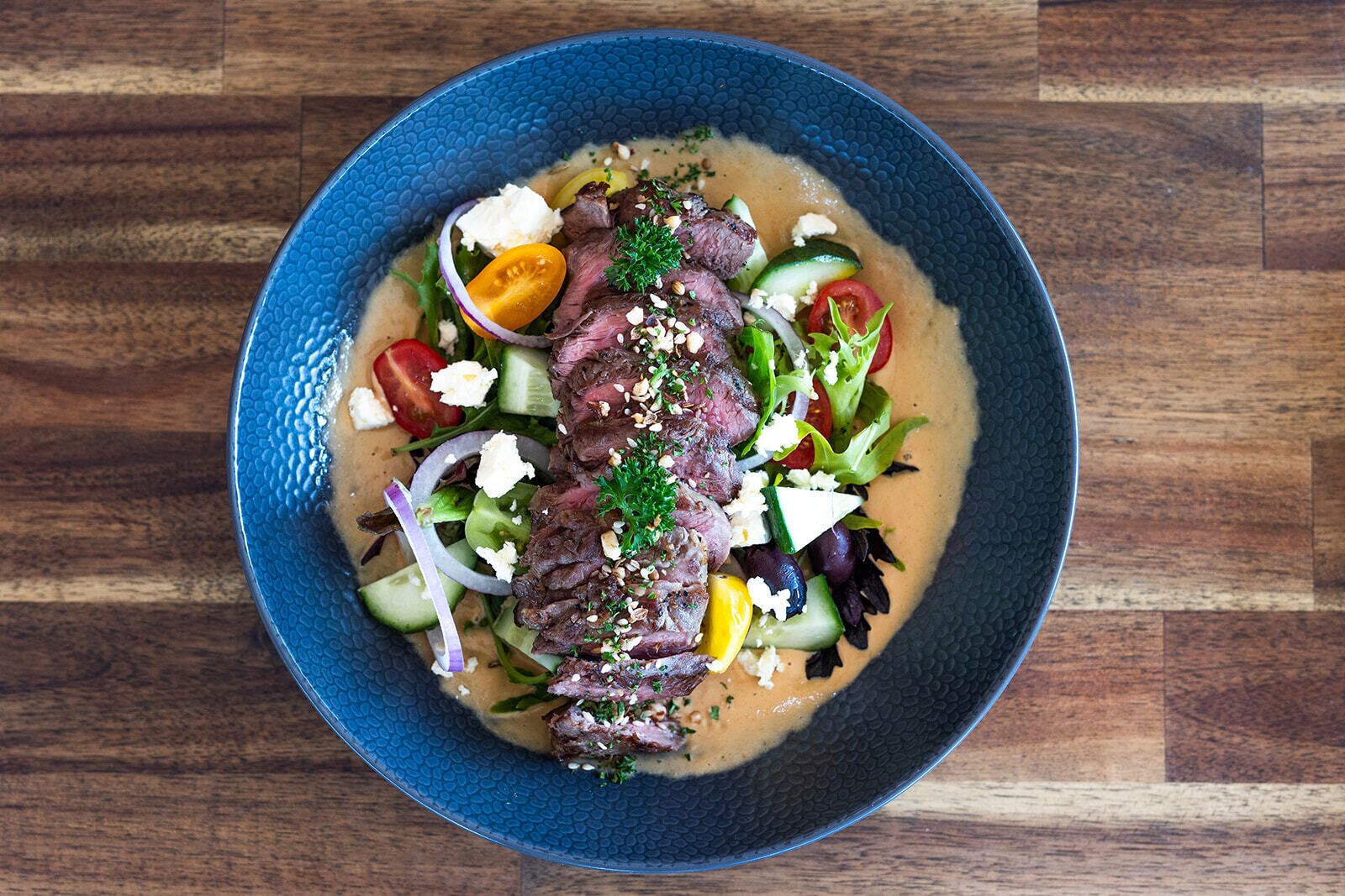 Um prato azul com bife, legumes e um molho cremoso sobre uma mesa de madeira.