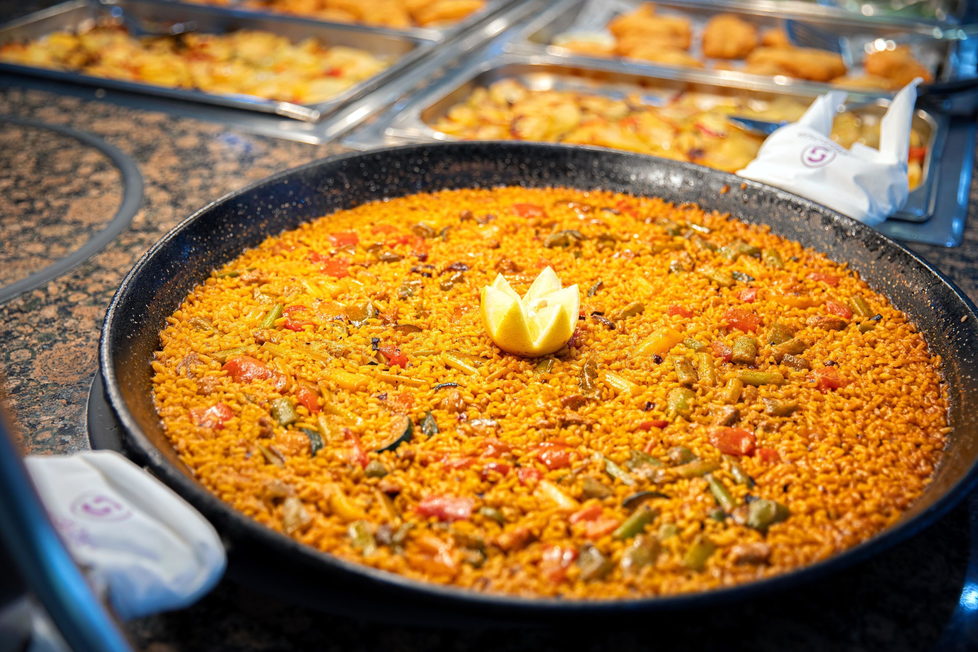 Uma travessa redonda e preta grande, cheia de um prato de arroz amarelo vibrante, decorado com uma fatia de limão, rodeada por diversos outros pratos em um buffet.