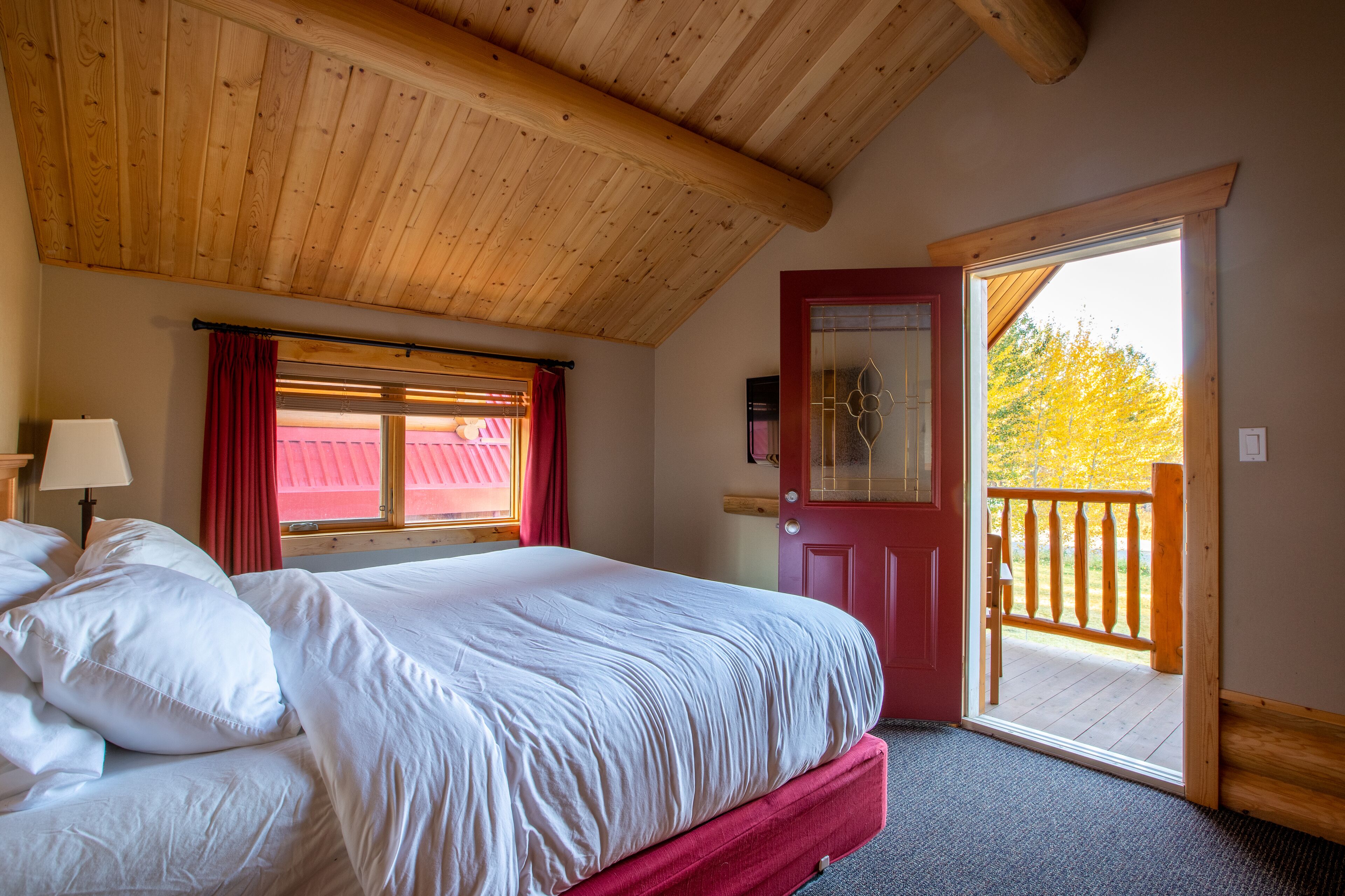 Quarto com teto de madeira, porta vermelha, janela com cortinas vermelhas e vista para um deck de madeira.