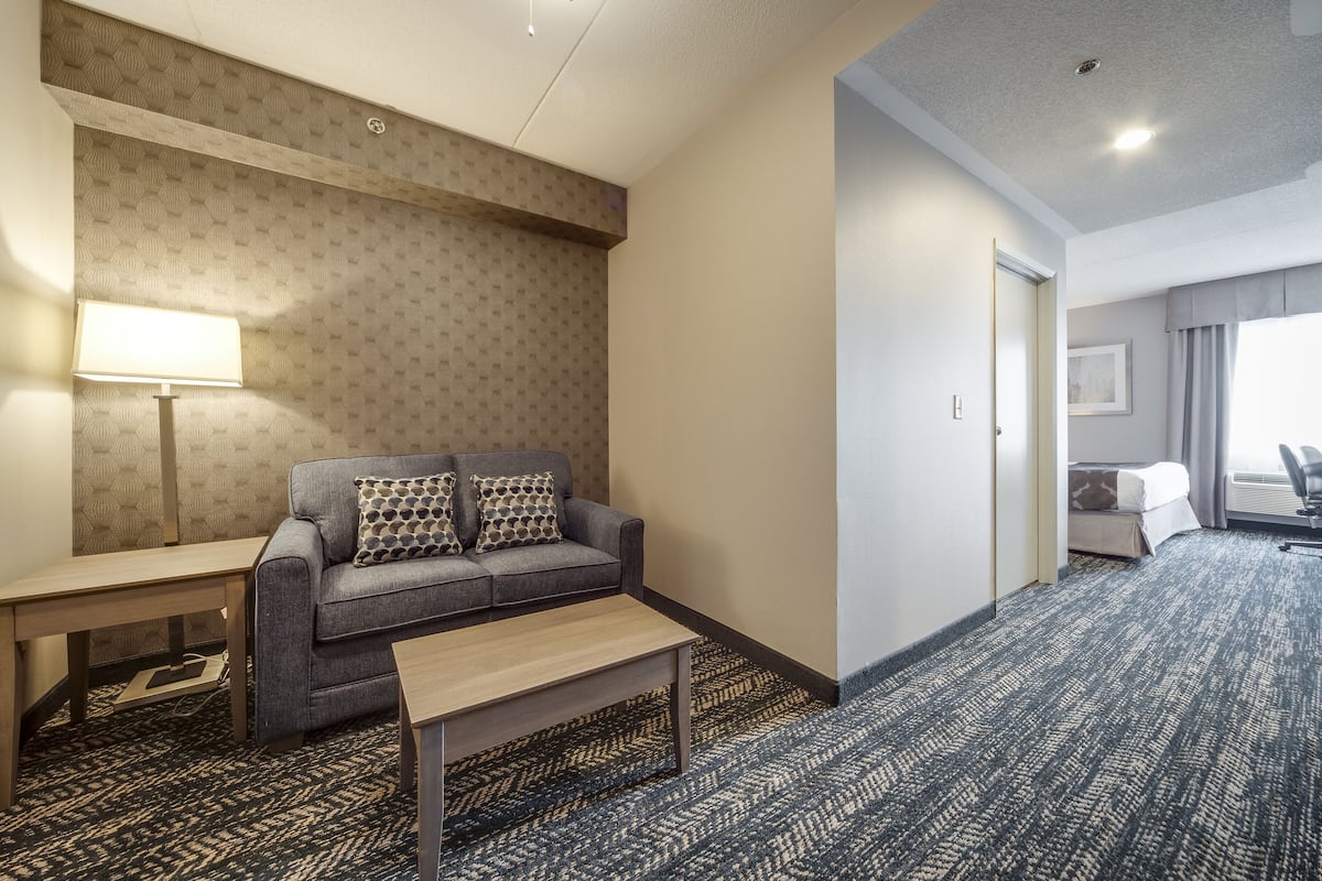 Quarto de hotel com sofá cinzento, mesa de centro de madeira, tapete estampado e cama com colcha cinza.