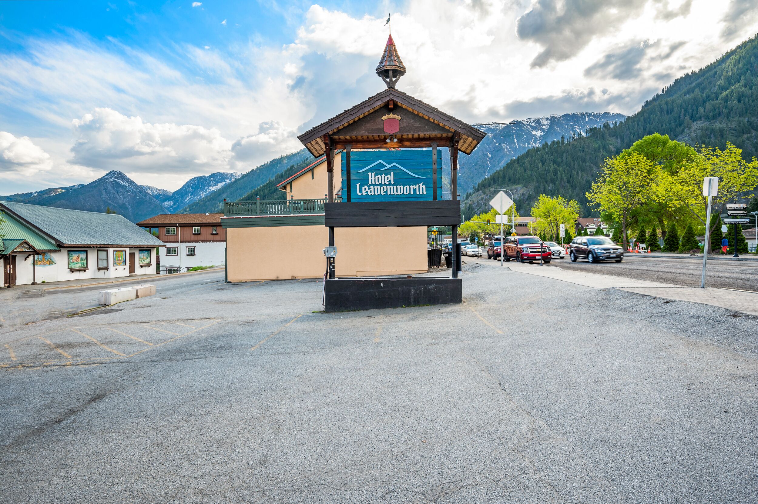 Entrada de um hotel com uma placa indicando "Hotel Leavenworth", rodeada por montanhas e um estacionamento.