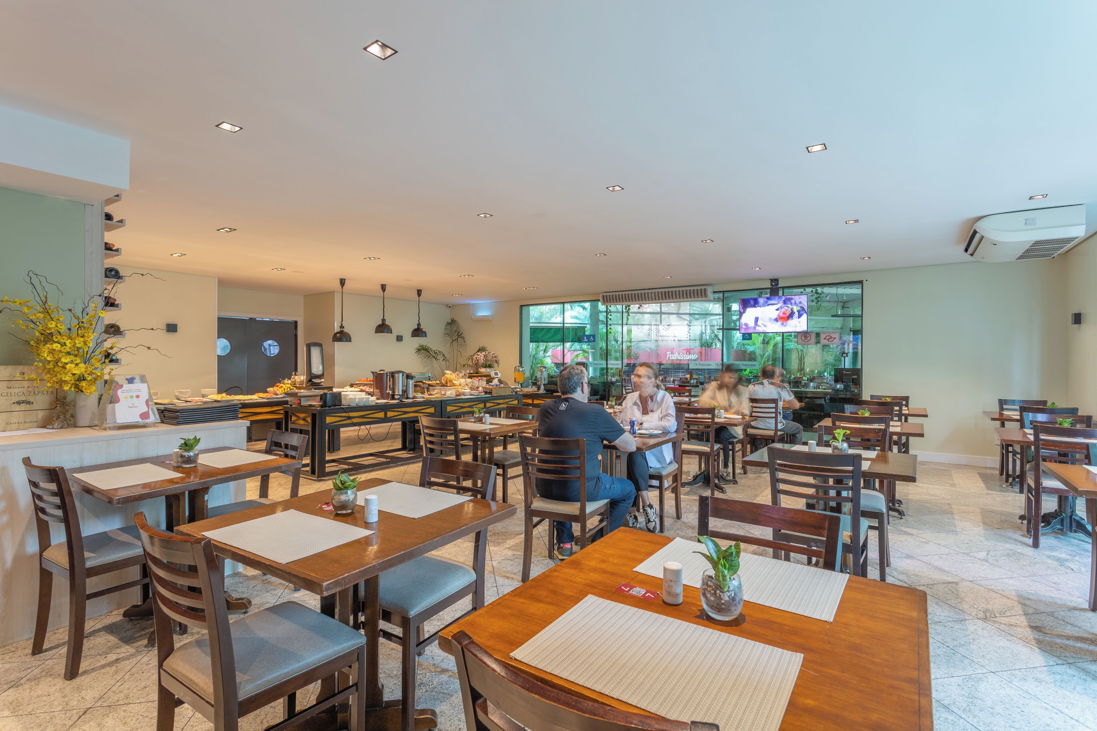 Um restaurante amplo com mesas e cadeiras de madeira, um balcão com cardápio e uma tela de televisão ao fundo.