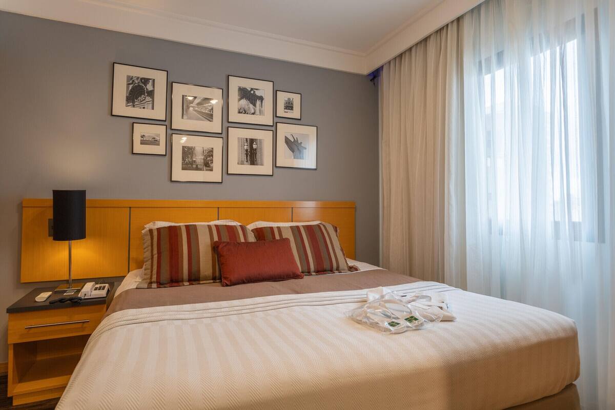 Quarto de hotel com uma cama, mesa de cabeceira com abajur, telefone e parede com quadros.