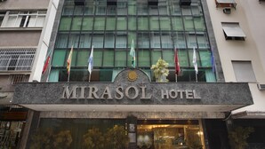 Um edifício de hotel alto com fachada moderna em vidro, várias bandeiras e uma entrada imponente com plantas em vasos.