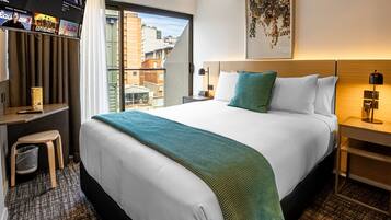 Quarto de hotel com uma cama grande, uma escrivaninha, uma cadeira, um cesto de lixo e uma janela com vista para a cidade.