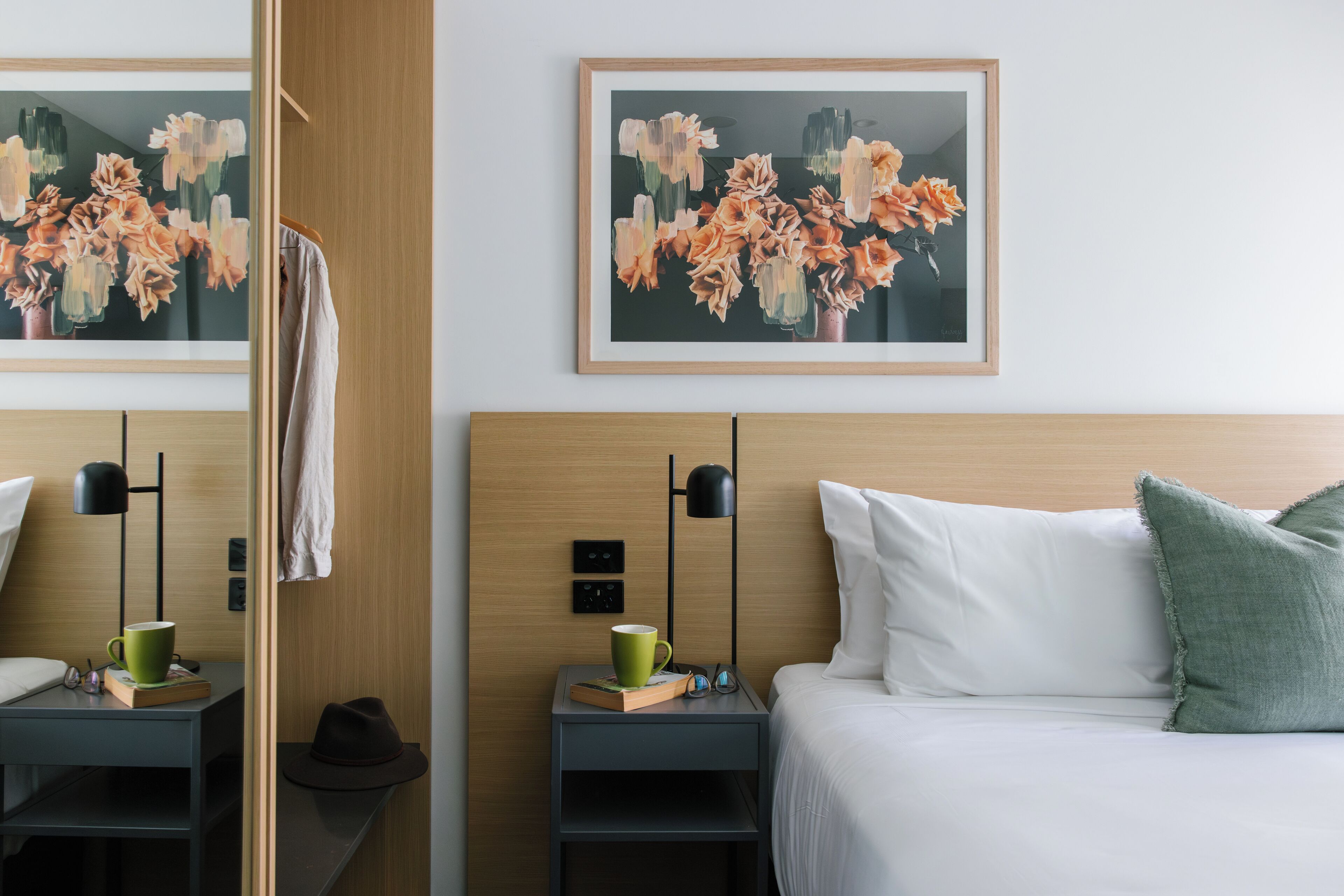 Quarto de hotel com uma cama, mesa de cabeceira com abajur, uma obra de arte floral emoldurada e um espelho refletindo o quarto.