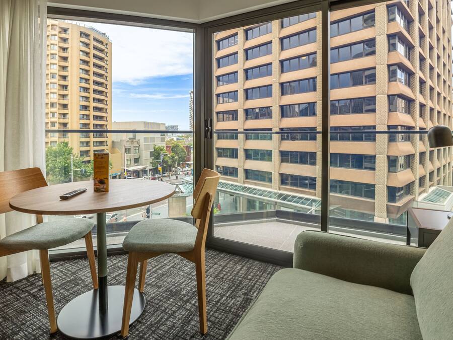 Quarto de hotel com varanda, mesa redonda com duas cadeiras, sofá e vista para edifícios altos.