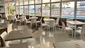 Uma área de refeições interna com mesas e cadeiras, grandes janelas oferecendo vista para o mar e palmeiras.