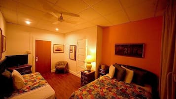 Quarto de hotel com duas camas, um ventilador de teto, uma escrivaninha, uma cadeira, um abajur e um quadro na parede.