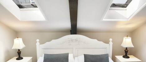 Quarto com cama branca, duas mesas de cabeceira com luminárias e claraboias.