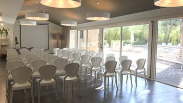 Sala de conferências com cadeiras brancas dispostas em fileiras, janelas amplas e iluminação pendente moderna.