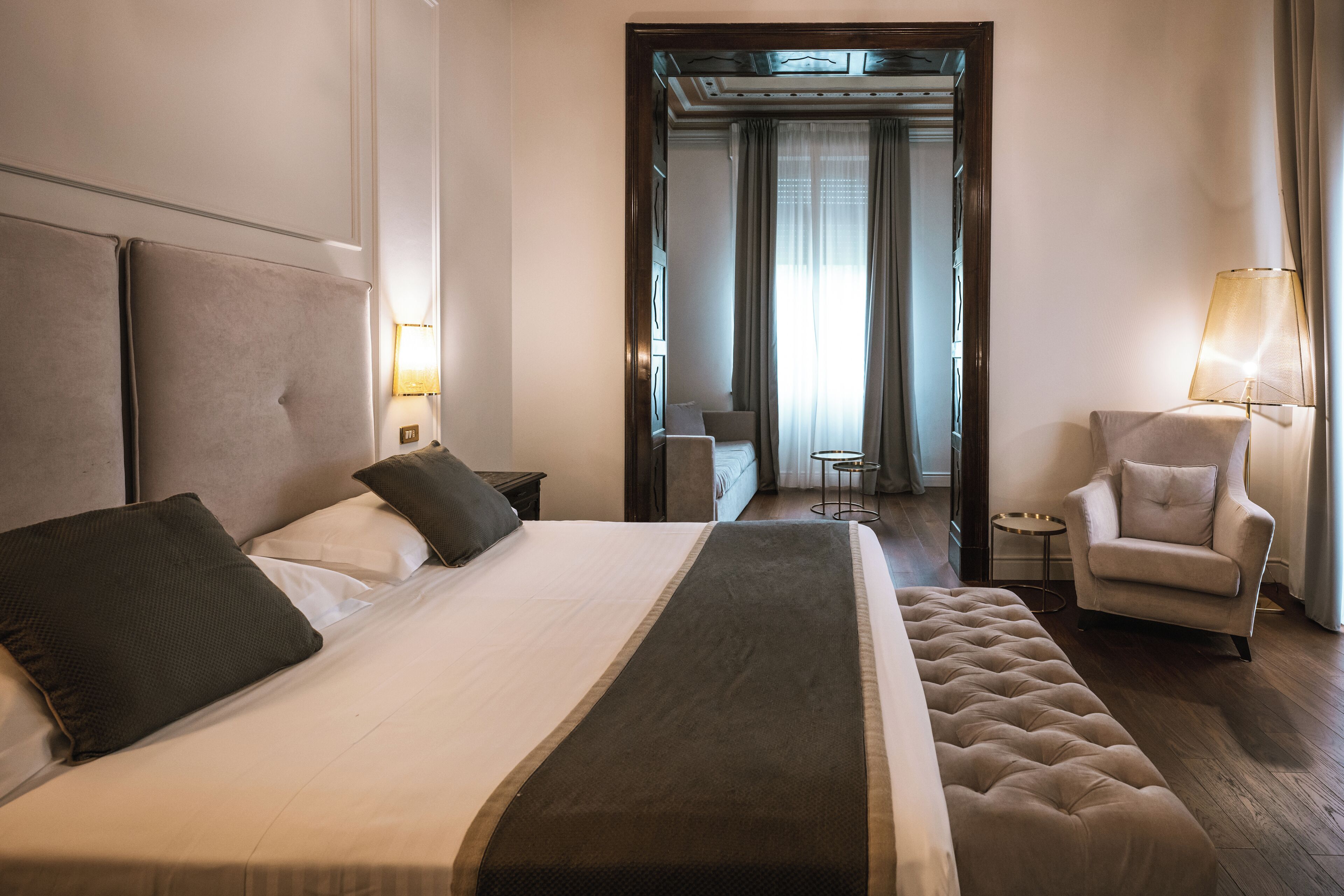 Quarto de hotel com uma cama grande, área de estar com uma poltrona, uma mesinha e uma luminária.