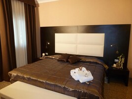 Quarto de hotel com uma cama grande, cabeceira, mesa de cabeceira e um banco.