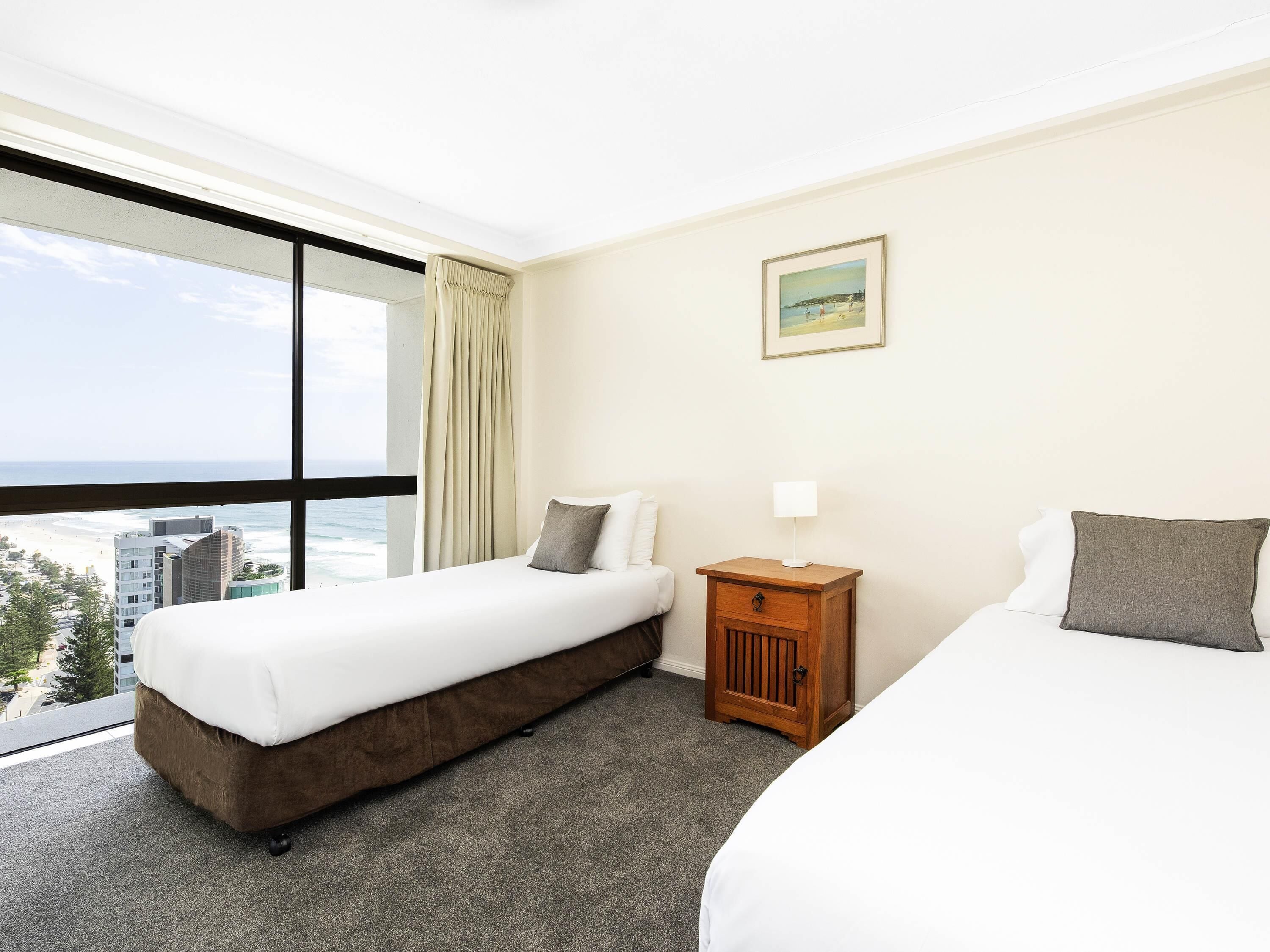 Quarto de hotel com duas camas, uma mesa de cabeceira, abajur, um quadro e vista para o mar.
