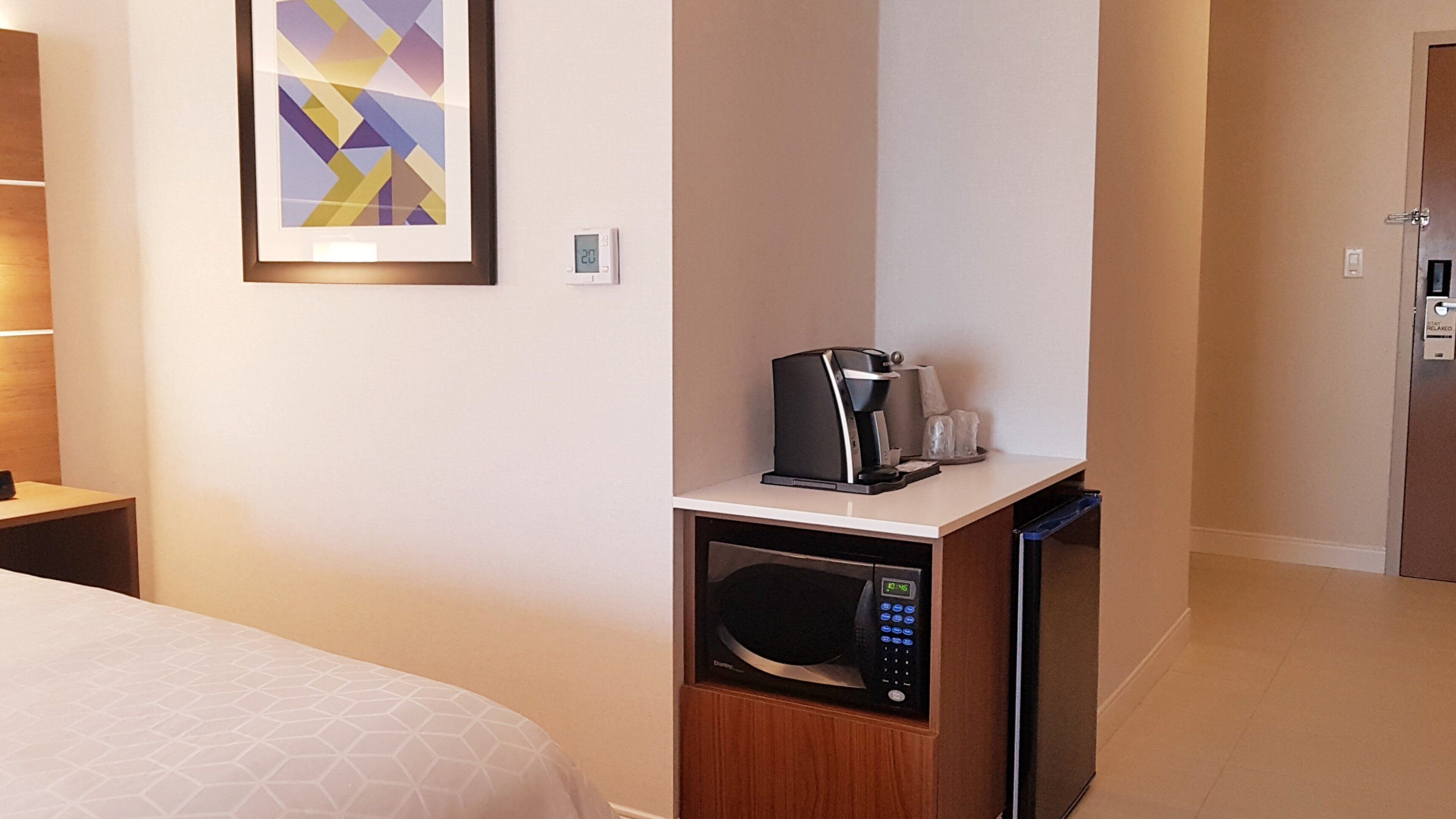 Quarto de hotel com cama, pequeno espaço de cozinha equipado com cafeteira e micro-ondas, e uma obra de arte abstrata emoldurada na parede.