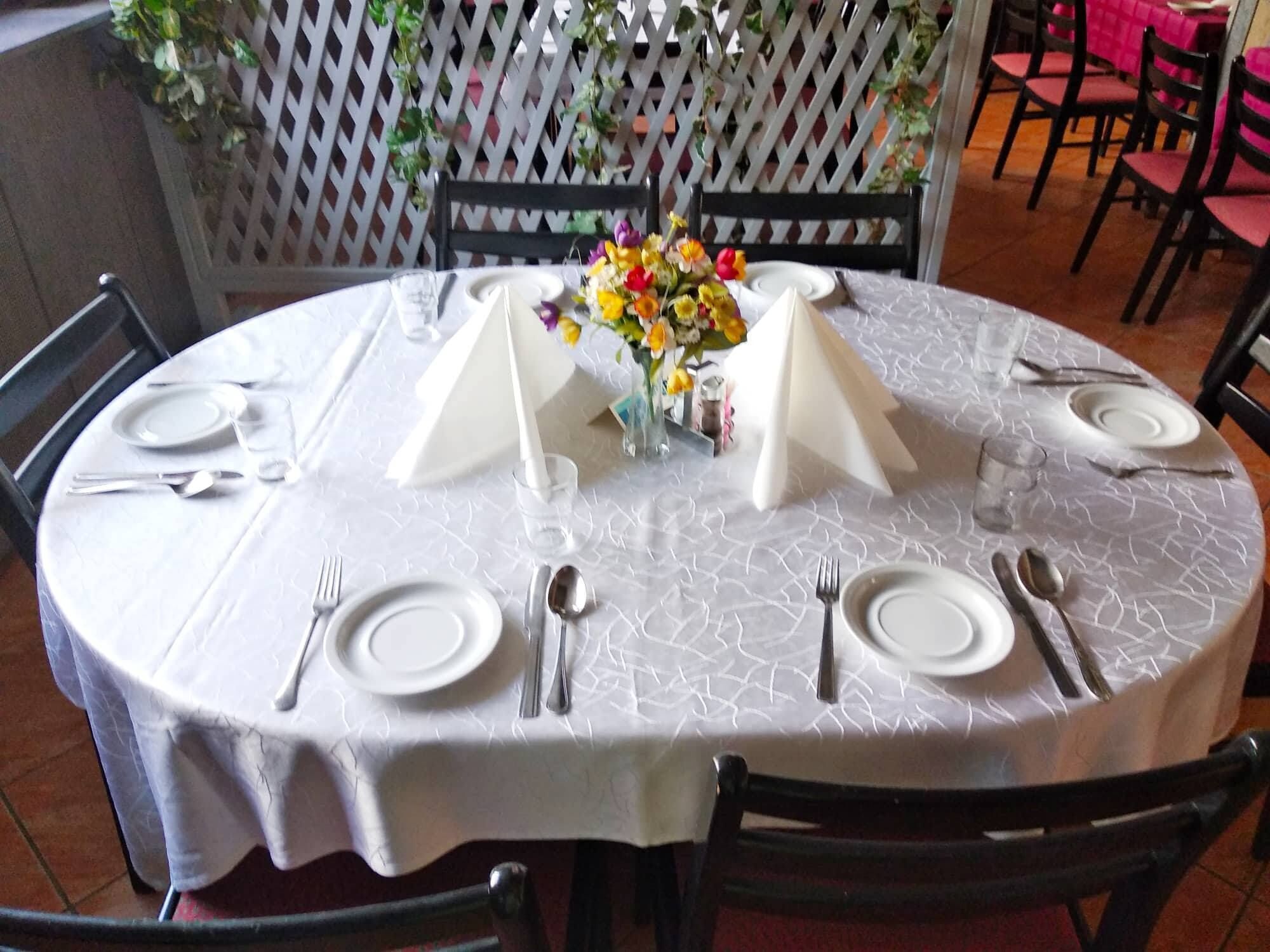 Uma mesa de jantar redonda com toalha branca, posta para quatro pessoas, com pratos, talheres e um arranjo de flores no centro.