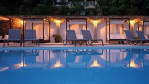 Área à beira da piscina com espreguiçadeiras e guarda-sóis, cercada por paredes de pedra e um edifício ao fundo.