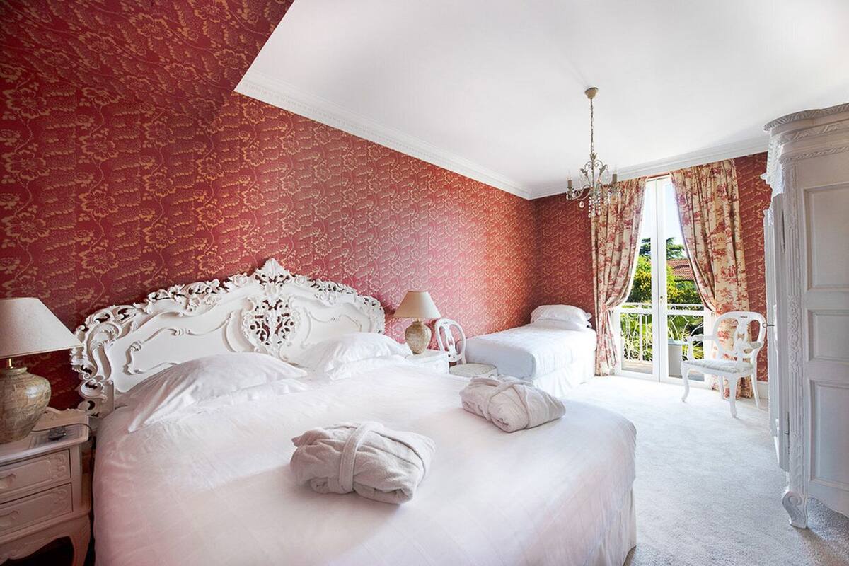 Quarto de hotel com uma cama grande, uma cama menor, um candelabro, uma janela com cortinas e vista para o verde lá fora.