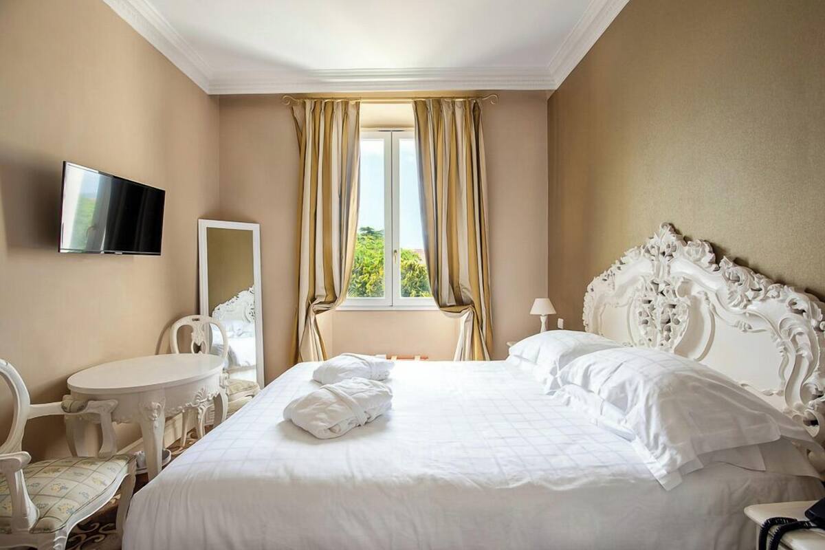 Quarto de hotel com uma cama grande, uma escrivaninha, uma cadeira, uma televisão e uma janela com cortinas.