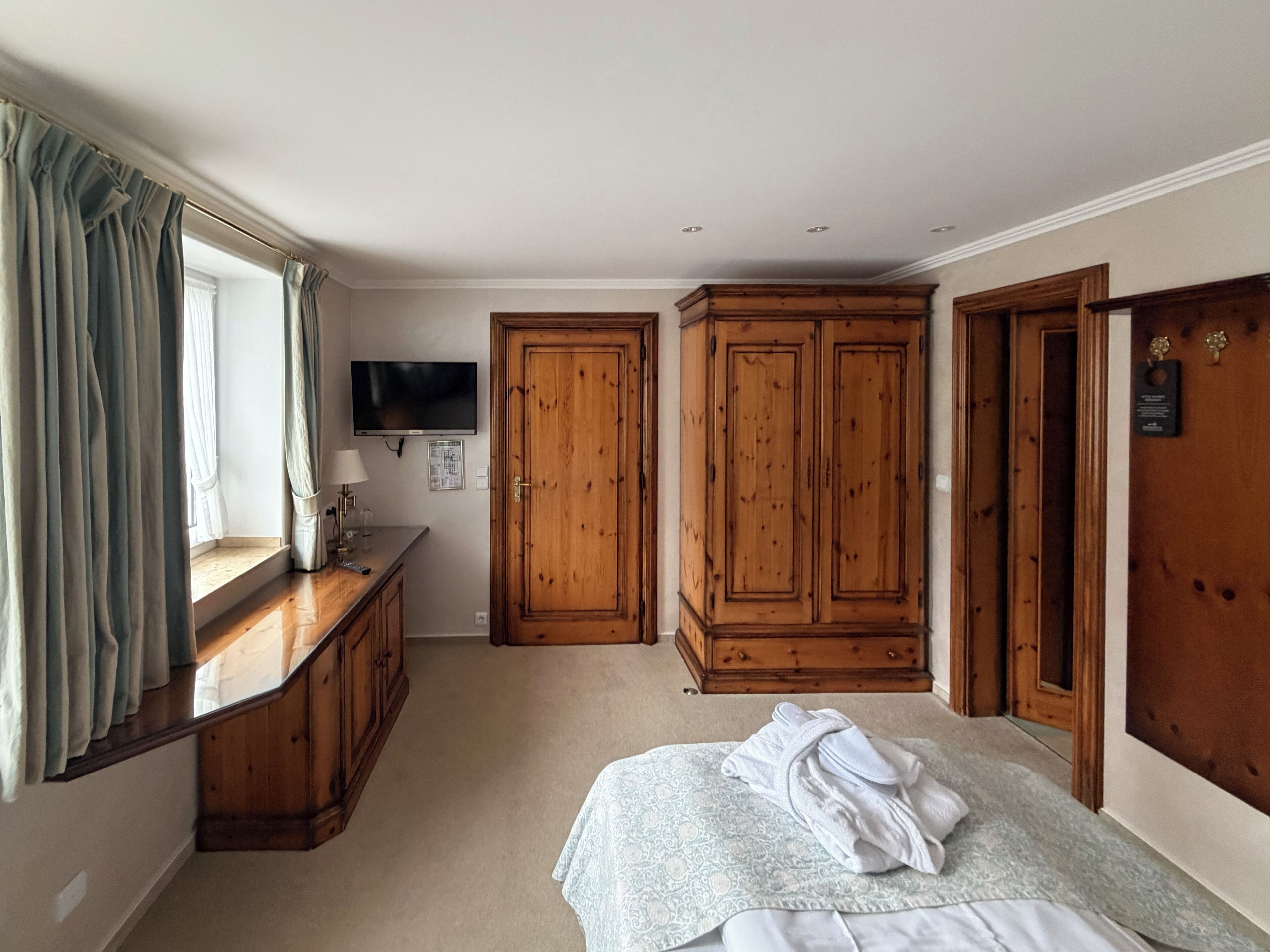 Quarto com cama, guarda-roupa de madeira, televisão e janela com cortinas.