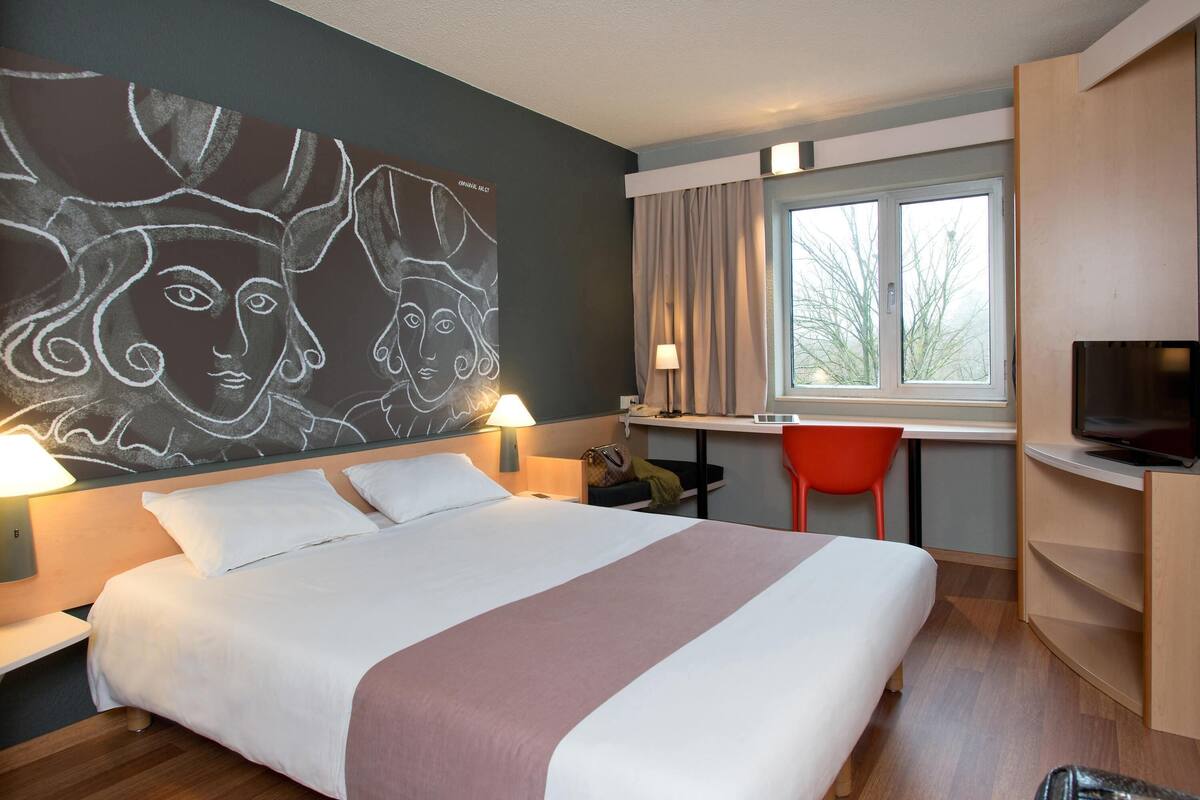 Quarto de hotel com uma cama, uma escrivaninha, uma cadeira, uma televisão e uma janela com cortinas.