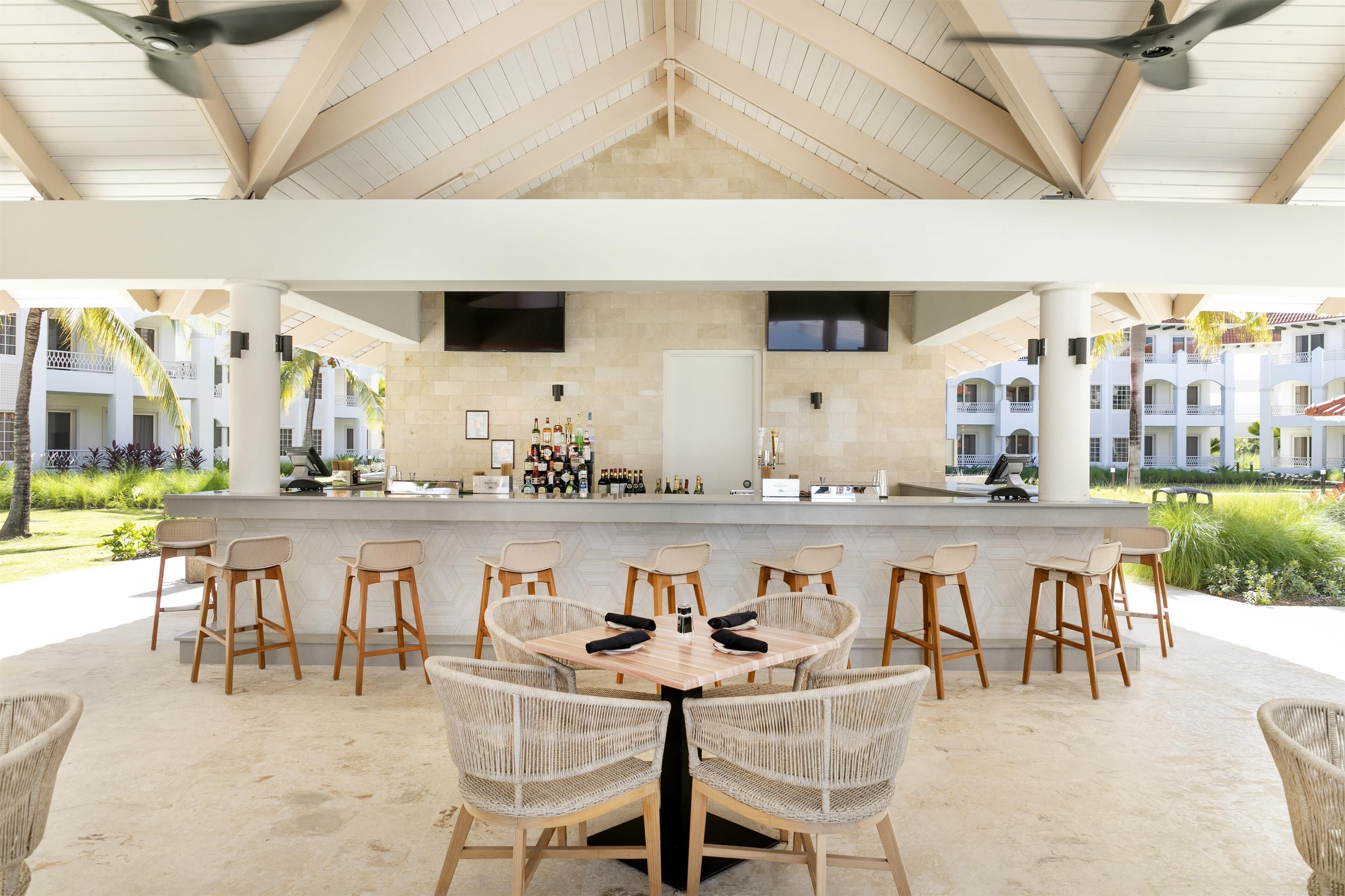 Uma área de bar ao ar livre com uma mesa redonda e seis banquetas, um ventilador de teto e vista para um edifício com varandas.