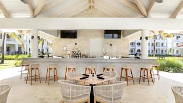 Uma área de bar ao ar livre com uma mesa redonda e seis banquetas, um ventilador de teto e vista para um edifício com varandas.