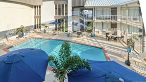 Área de piscina com espreguiçadeiras e guarda-sóis, um edifício com varandas, e um céu claro.
