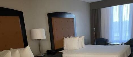 Quarto de hotel com duas camas, mesa de cabeceira, luminária e janela com cortinas.