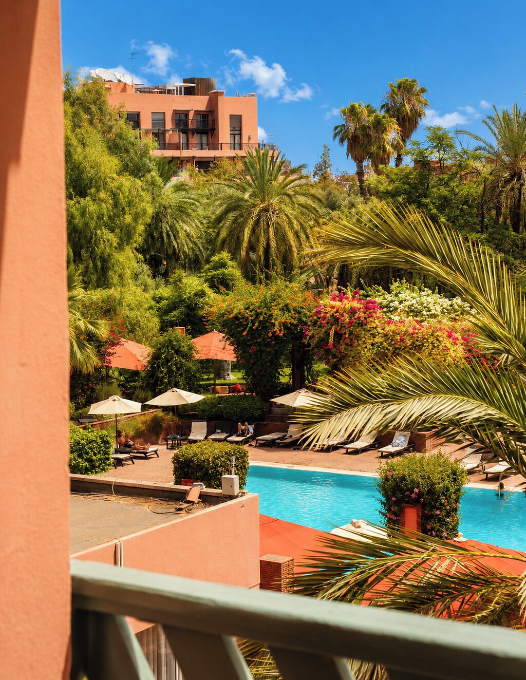 Área de piscina de hotel com espreguiçadeiras e guarda-sóis, rodeada por vegetação exuberante e um céu azul límpido.