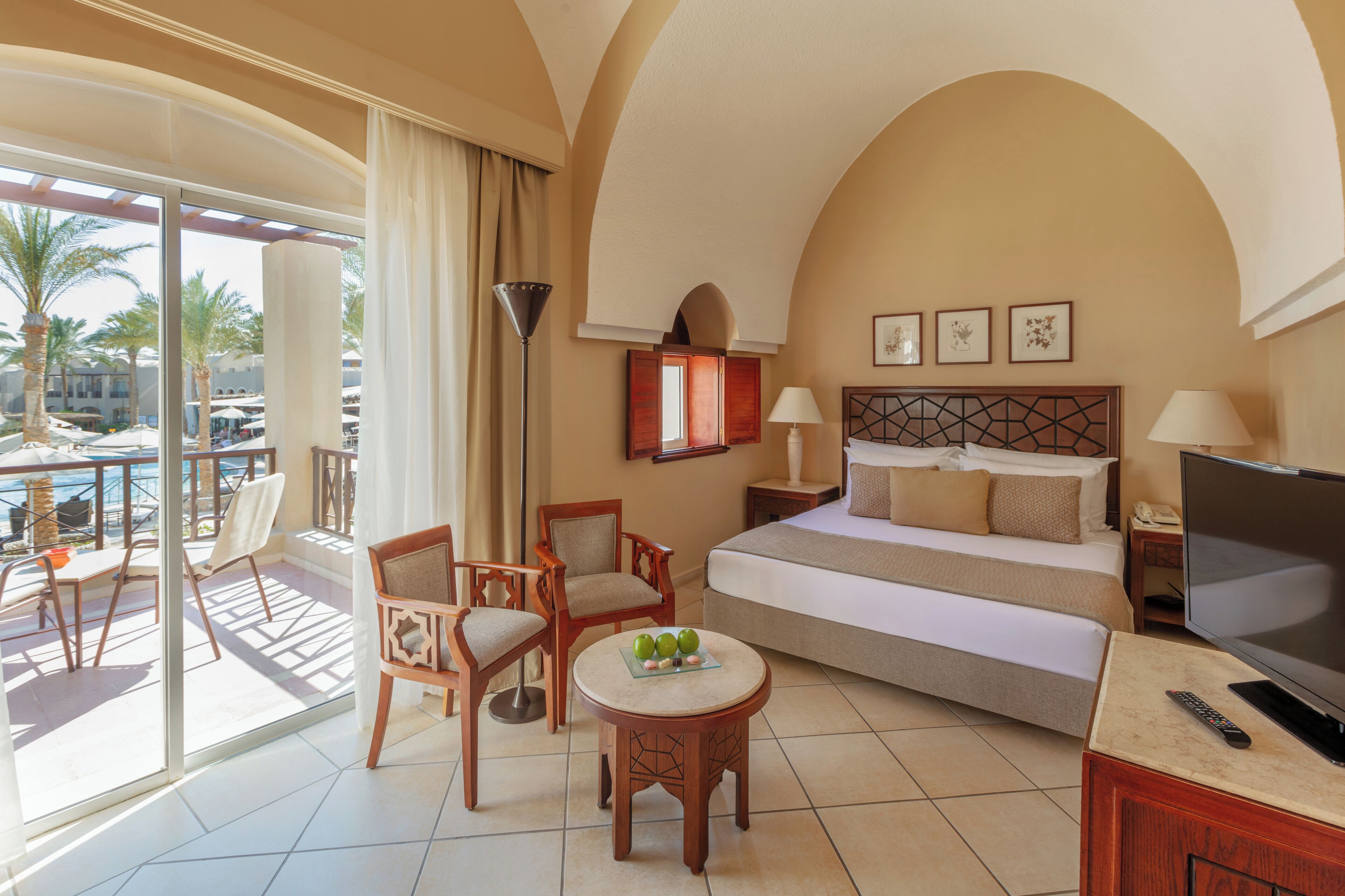 Quarto de hotel com varanda, uma cama, duas cadeiras, uma mesinha com frutas e uma televisão.