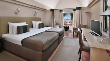 Quarto de hotel com uma cama grande, uma escrivaninha, uma cadeira e uma TV. Possui uma varanda com vista para o mar.