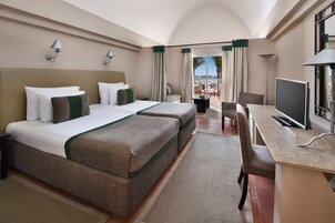 Quarto de hotel com uma cama grande, uma escrivaninha, uma cadeira e uma TV. Possui uma varanda com vista para o mar.