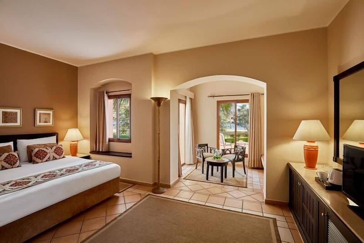 Quarto de hotel com uma cama, uma escrivaninha, uma cadeira e uma televisão. Há duas janelas com cortinas e uma porta que dá para uma varanda.