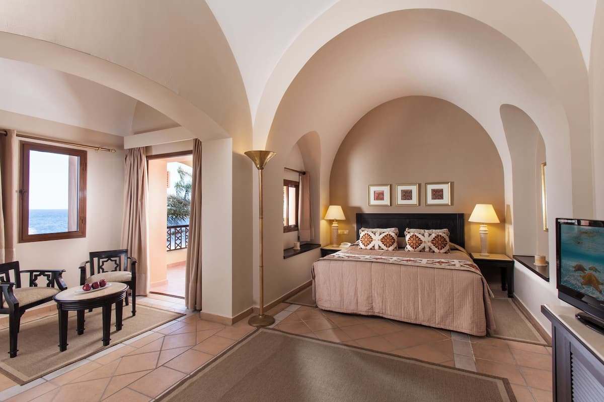Quarto de hotel com cama, televisão, escrivaninha com cadeira, luminária e vista para o mar através de uma varanda.