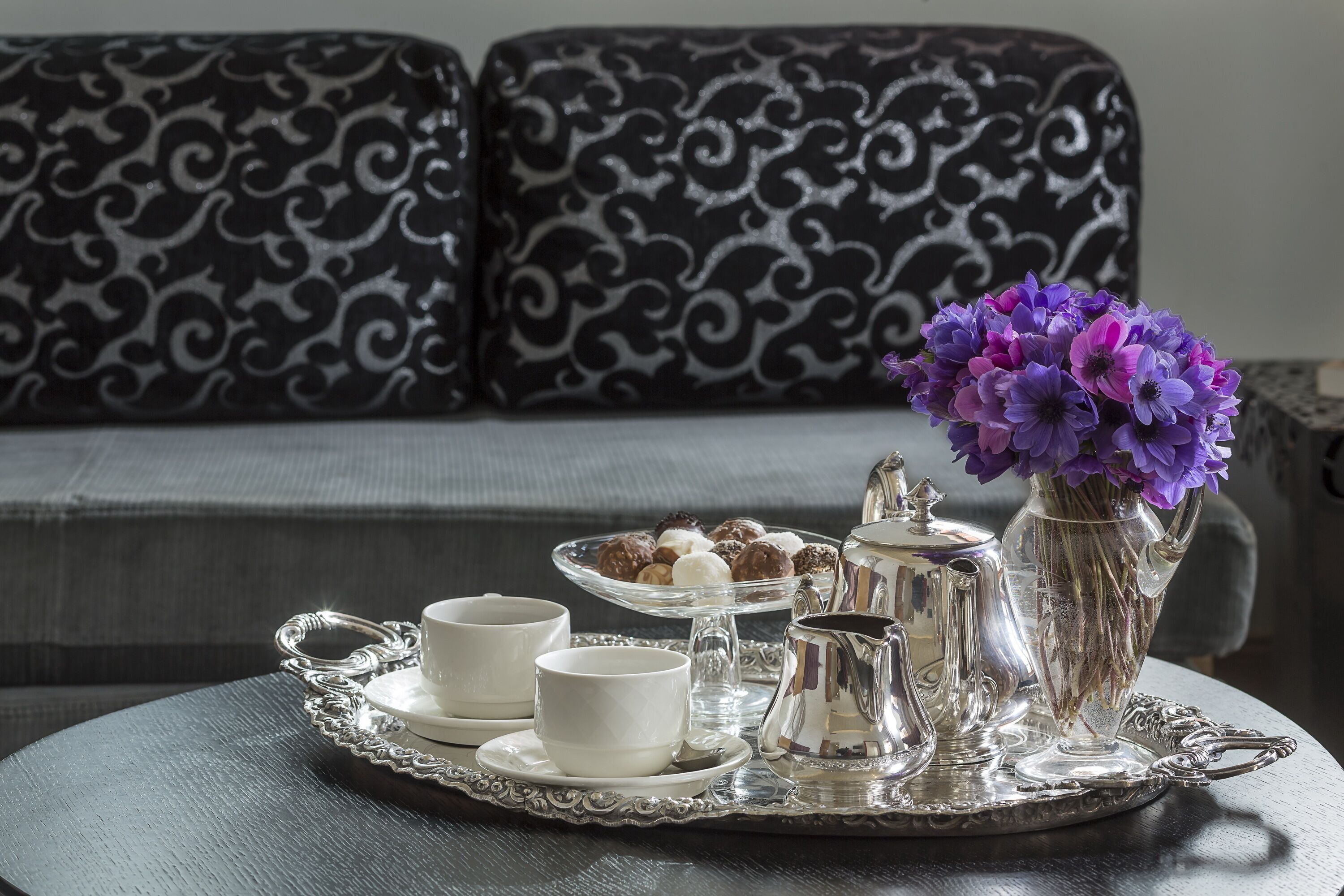 Uma bandeja com talheres, xícaras e um vaso de flores roxas sobre uma mesa redonda.