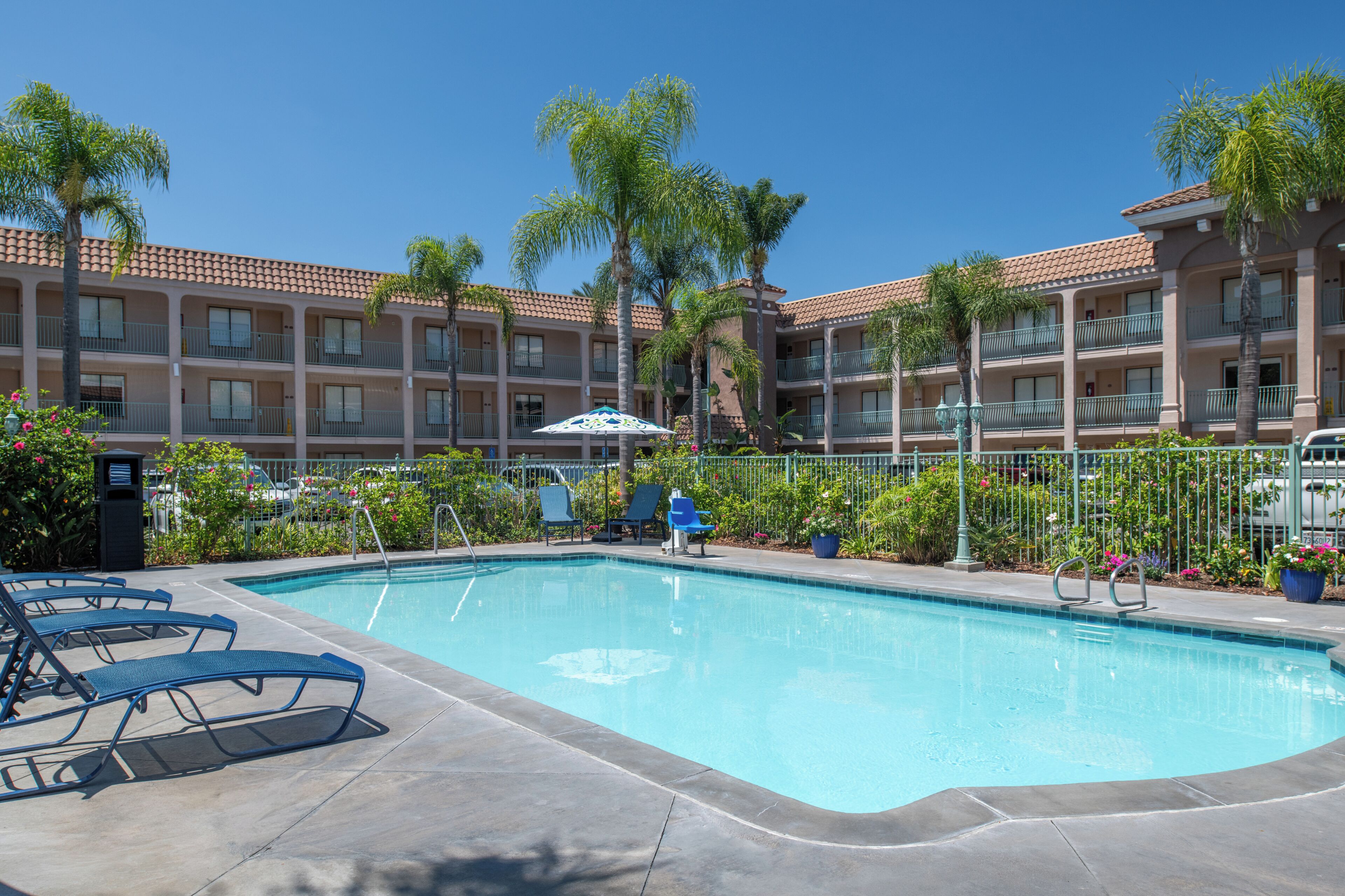 Área de piscina de hotel com piscina azul cristalina, espreguiçadeiras e um edifício com varandas ao fundo.