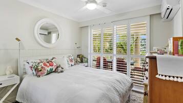 Quarto com cama, espelho redondo, ventilador de teto e janela com persianas.