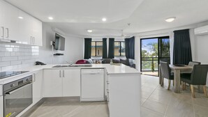 Uma cozinha moderna com armários brancos, forno embutido e máquina de lavar louça. Há uma área de refeições com uma mesa e cadeiras de madeira.