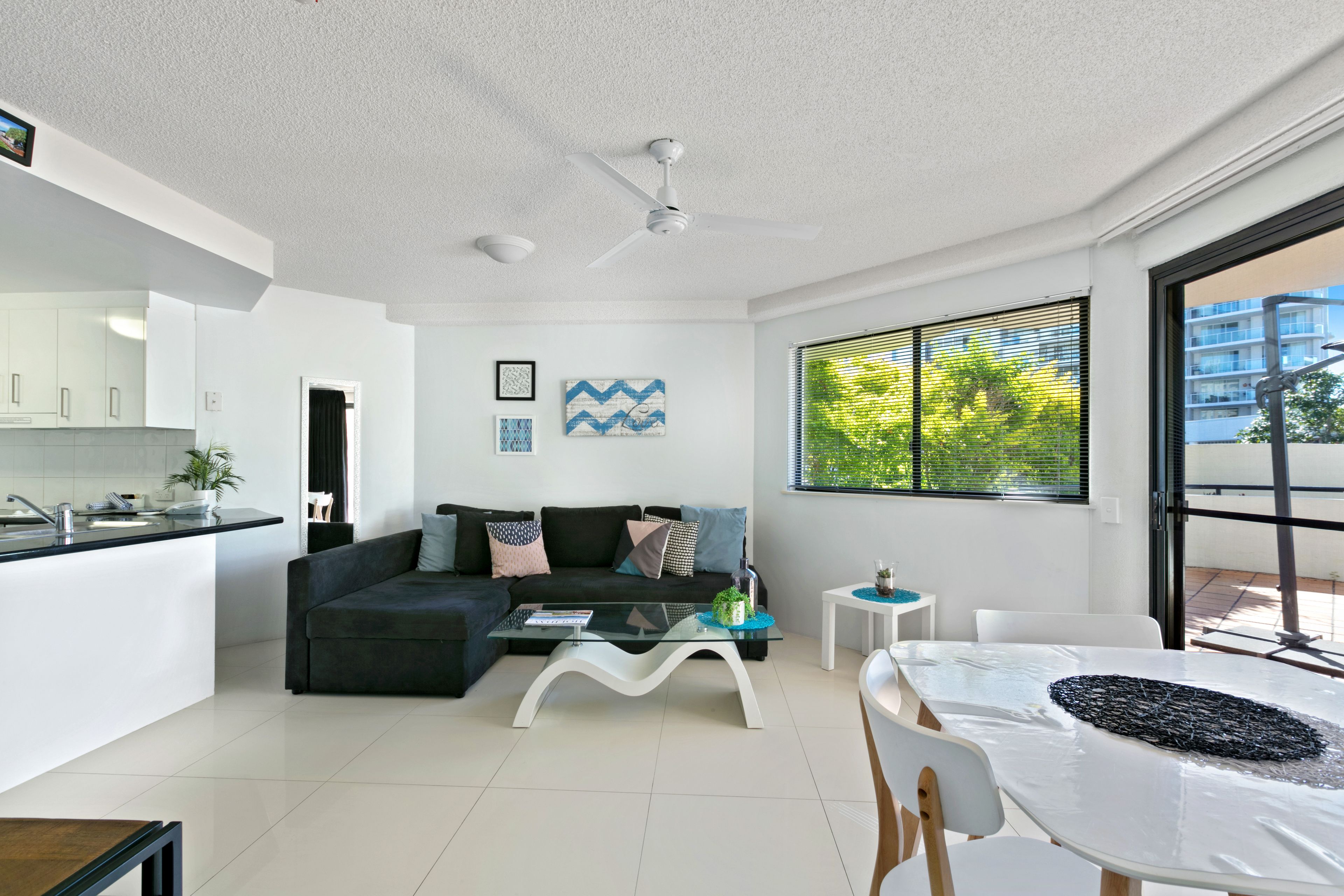 Uma sala de estar moderna com um sofá escuro, uma mesa de centro de vidro e um espaço de jantar com uma mesa e cadeiras brancas.