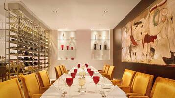 Uma sala de jantar com uma mesa comprida posta para uma refeição, rodeada por cadeiras douradas, e uma adega à esquerda.
