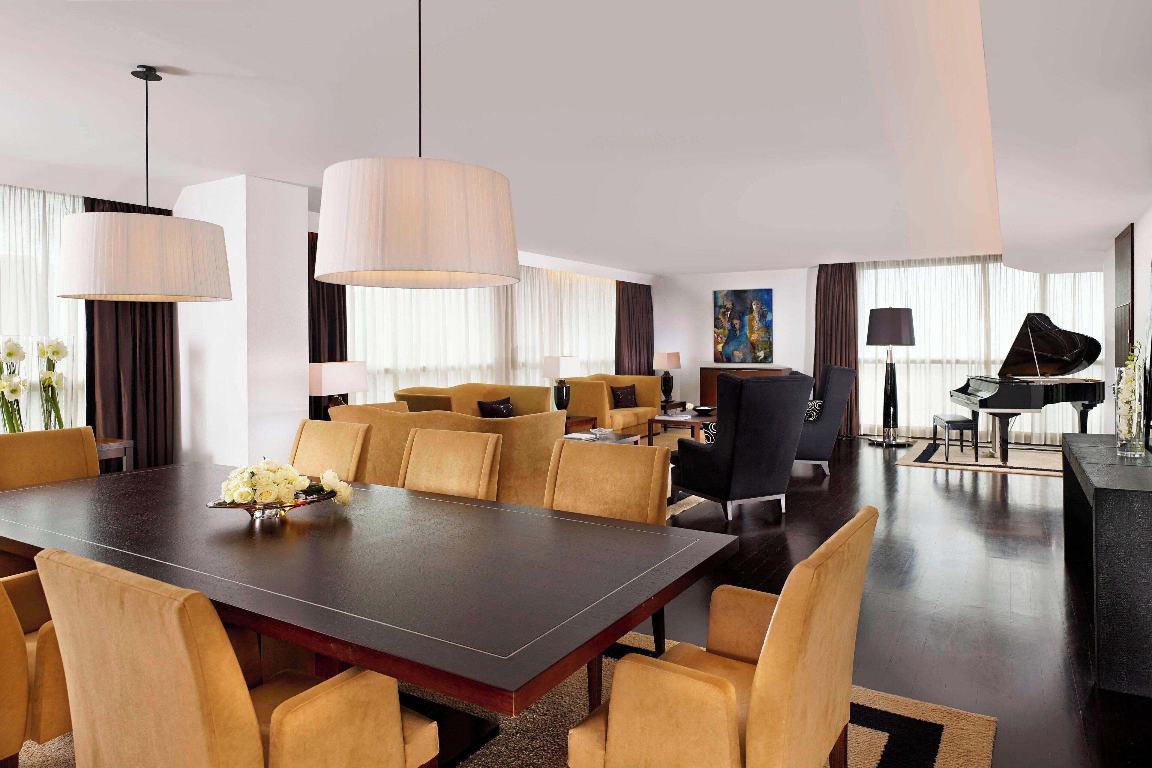 Quarto de hotel moderno com uma grande mesa de jantar, assentos confortáveis, um piano e uma janela ampla com cortinas.