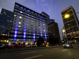 Um hotel com vários andares, com luzes azuis iluminadas na fachada, um estacionamento com vários carros e uma rua com um veículo em movimento.