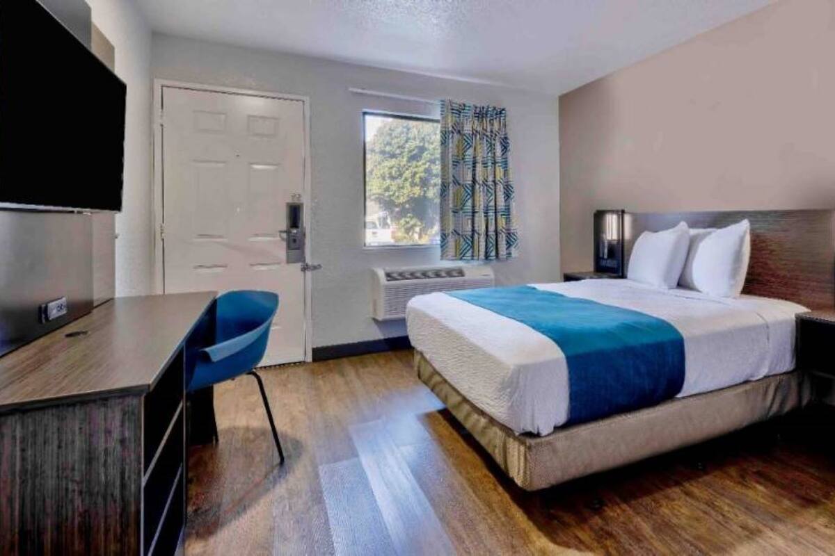 Quarto de hotel com cama, escrivaninha, cadeira, televisão e janela com cortinas.
