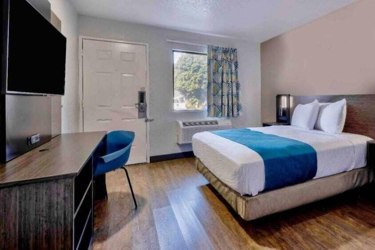 Quarto de hotel com cama, escrivaninha, cadeira, televisão e janela com cortinas.